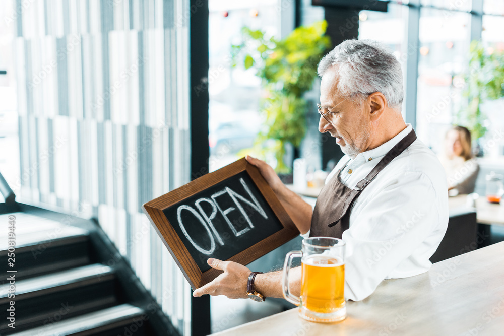 senior male owner of pub holding open sign and standing near bar ...
