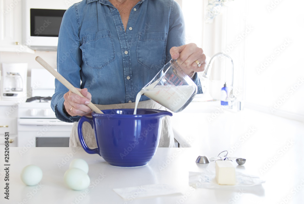 Photograph of a woman in the kitchen pouring milk into a mixing bowl ...