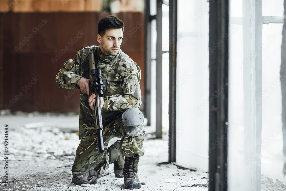 A young military soldier sits with a big rifle in his hands, near the ...