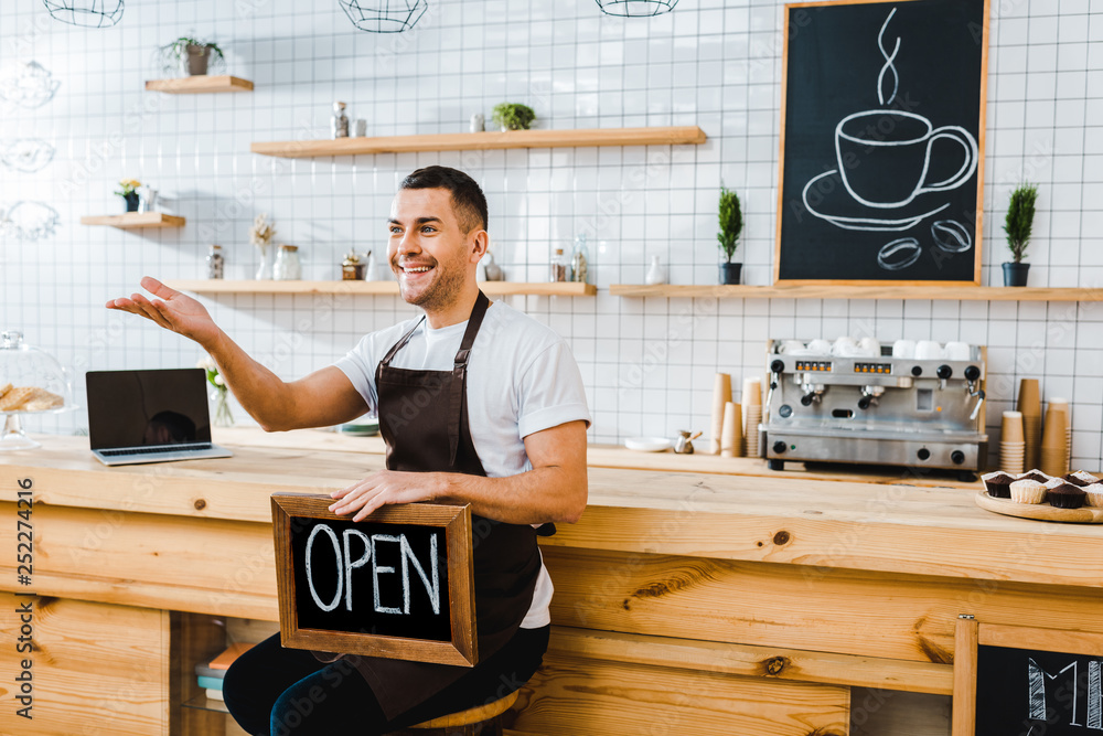 handsome cashier in apron sitting near wooden bar counter, holding ...