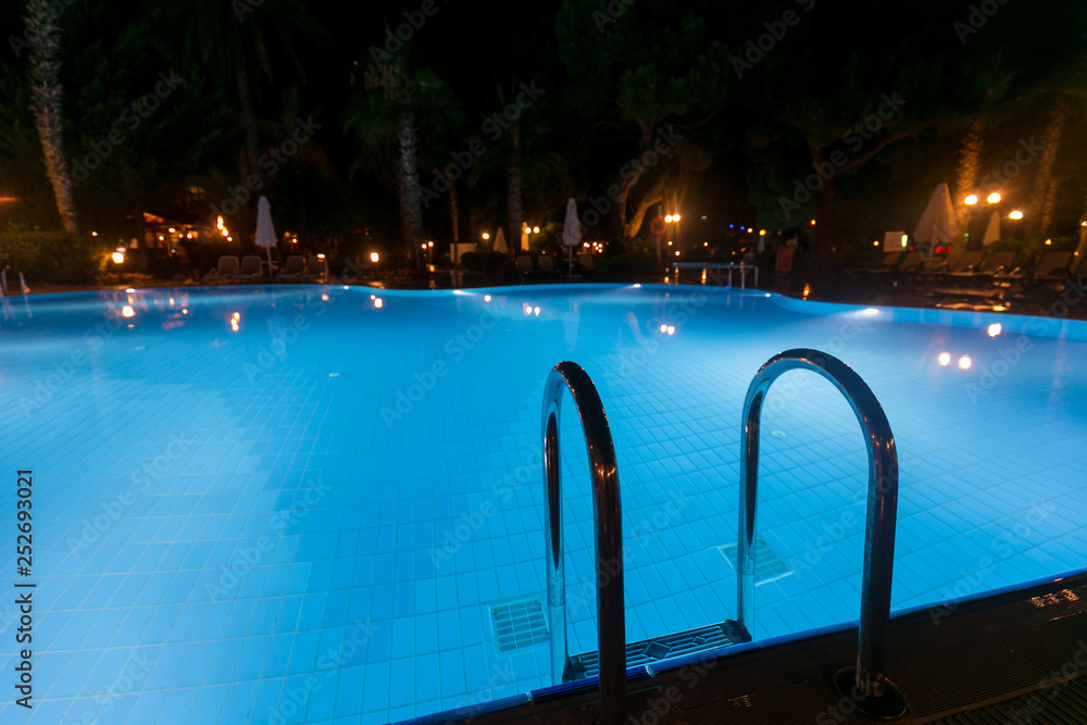 Illuminated swimming pool at night time. Grab bars ladder in the blue ...