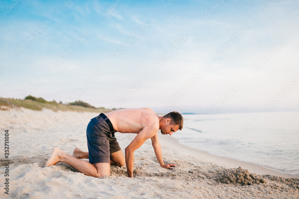 Young topless barefoot man in shorts digging hole by hands on beach ...