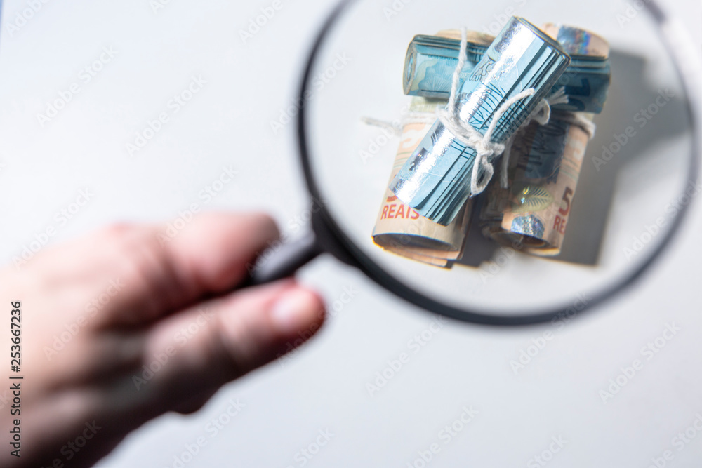 Out of focus hand with magnifying glass looking at rolls of banknotes ...
