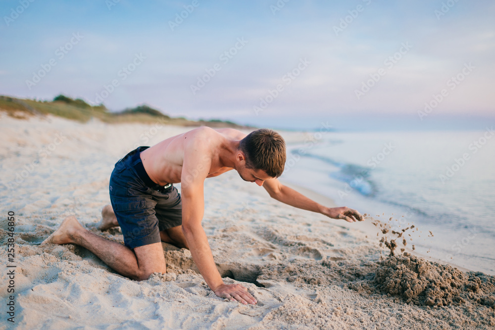 Young topless barefoot man in shorts digging hole by hands on beach ...