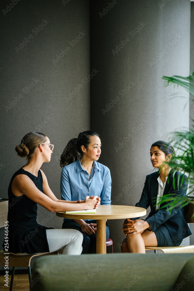 A diverse group of three young women sit around a table and smile while ...