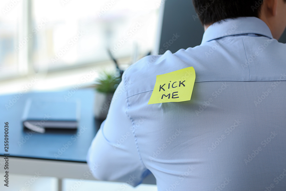 Sticky note with text KICK ME on back of man working in office. April ...