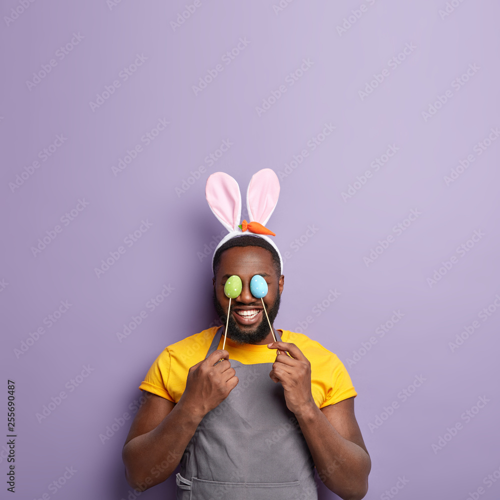 Glad African American man covers eyes with two Easter eggs, wears bunny ...