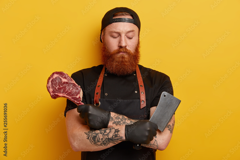 Serious red haired butcher holds raw fresh meat and cleaver, crosses ...