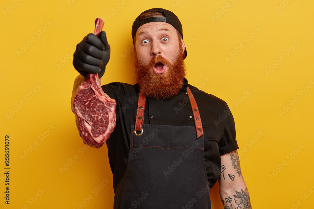 Foto de Surprised cheerful male butcher holds fresh raw piece of meat ...