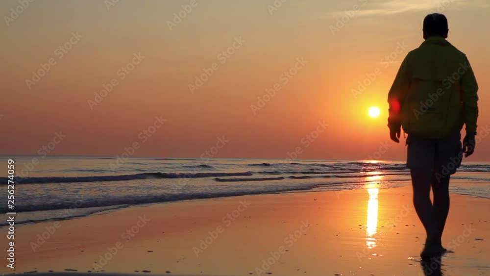 Man walking along a beach by a tranquil golden sunrise over the shore ...