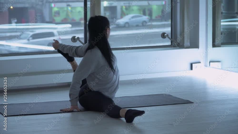 Young woman stratching legs on mat in the gym with large floor-to ...