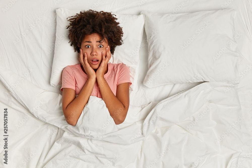 Top view of dark skinned woman scared of nightmare, lies in bed with ...