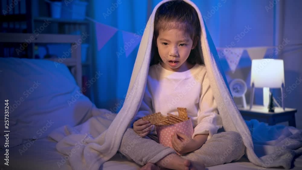 Happy child girl secretly eating candy under the blanket on bed in a ...