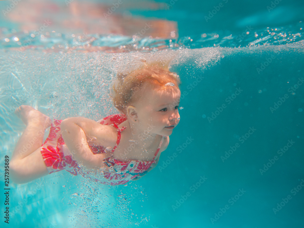 Smiling baby girl in cute modern dress diving underwater in blue ...