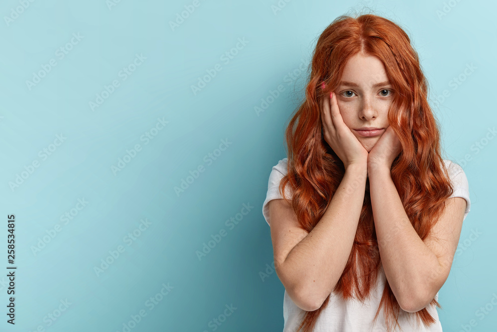 Indoor shot of sad lonely ginger girl touches cheeks with both hands ...