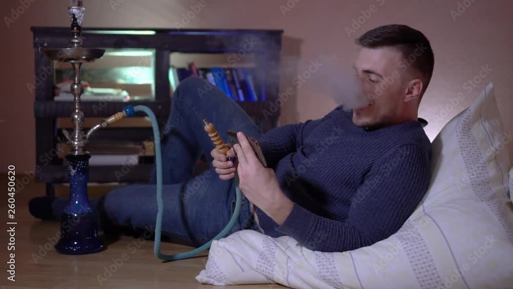 Vidéo Stock A young guy smokes a hookah, exhales smoke and watches the ...