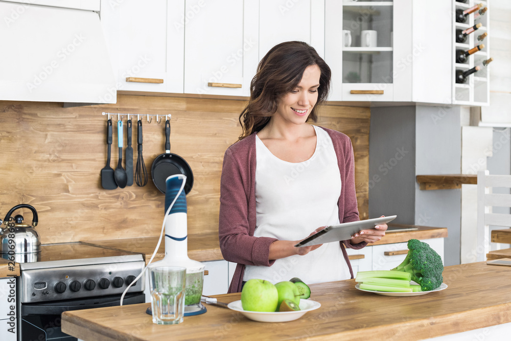 Young woman in kitchen following recipe on digital tablet and cooking ...