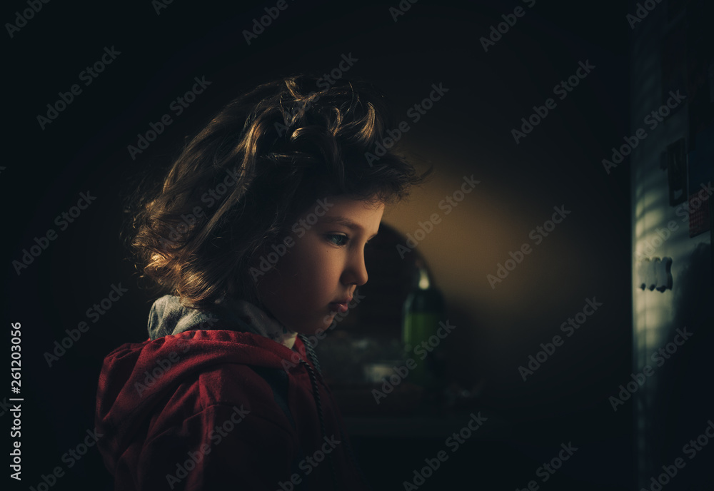 A cute little girl is standing in a dark room and looking away ...