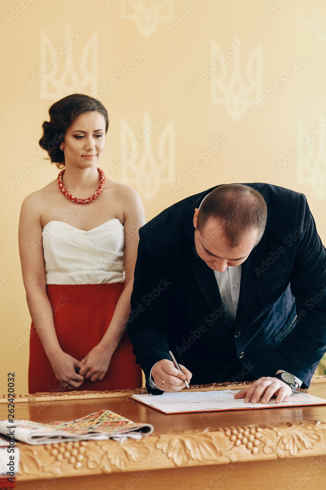 Handsome smiling groom in stylish suit signing marriage contract during ...