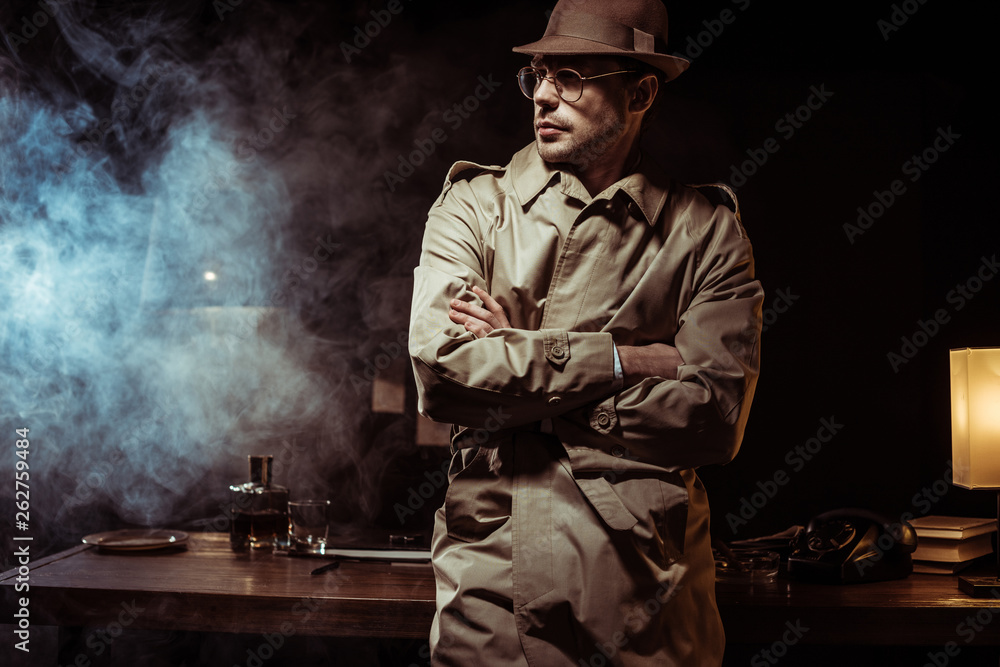Detective in trench coat and hat standing with crossed arms in office ...