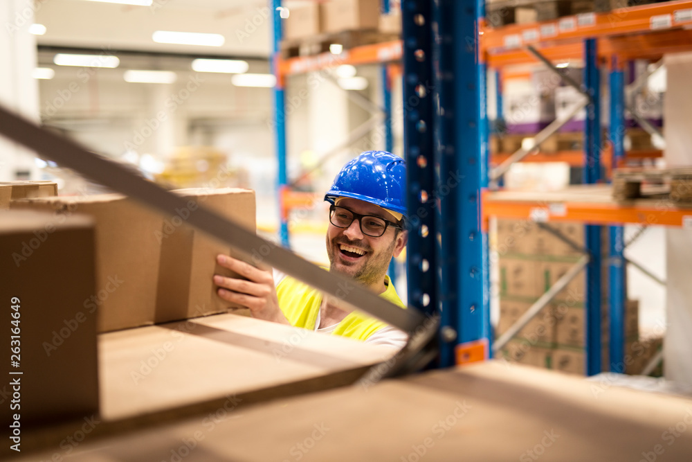 Warehouse worker picking up boxes in large warehouse distribution center. Man with hardhat and ...