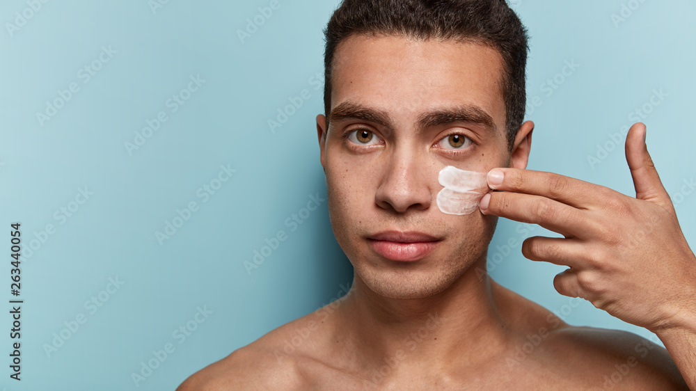 Serious male model with muscular body, applies face cream, cares of ...