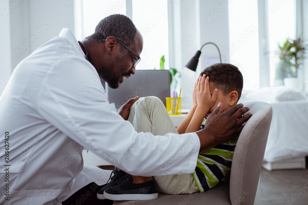 Dark-skinned father working as doctor talking to son hiding his face ...