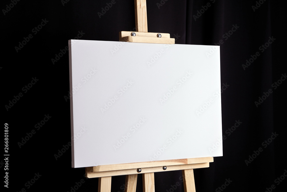 White blank canvas stands on a wooden artistic easel on black curtain ...