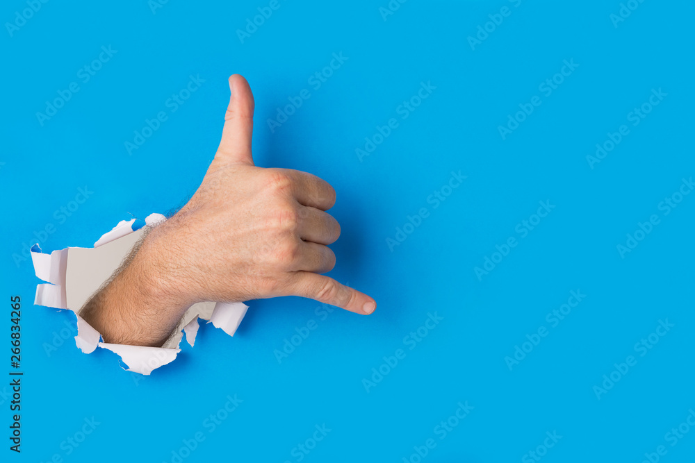Male hand tearing through blue paper background creating a shaka hand ...