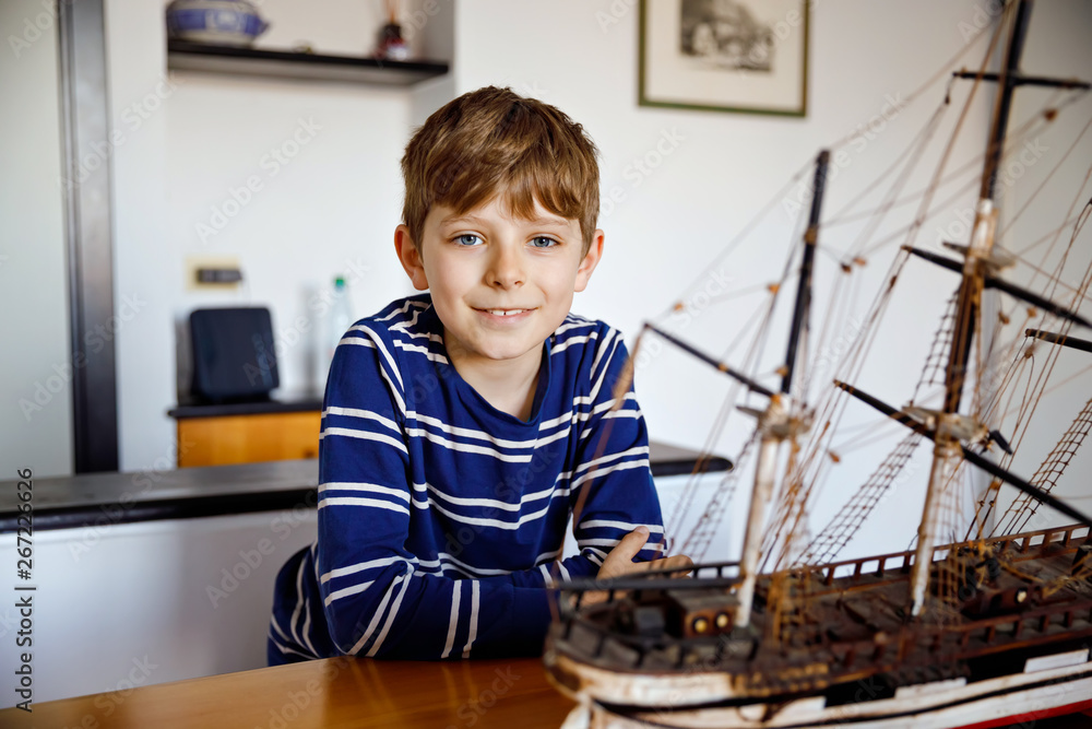 Little blond school kid boy playing with sailing ship model indoors ...