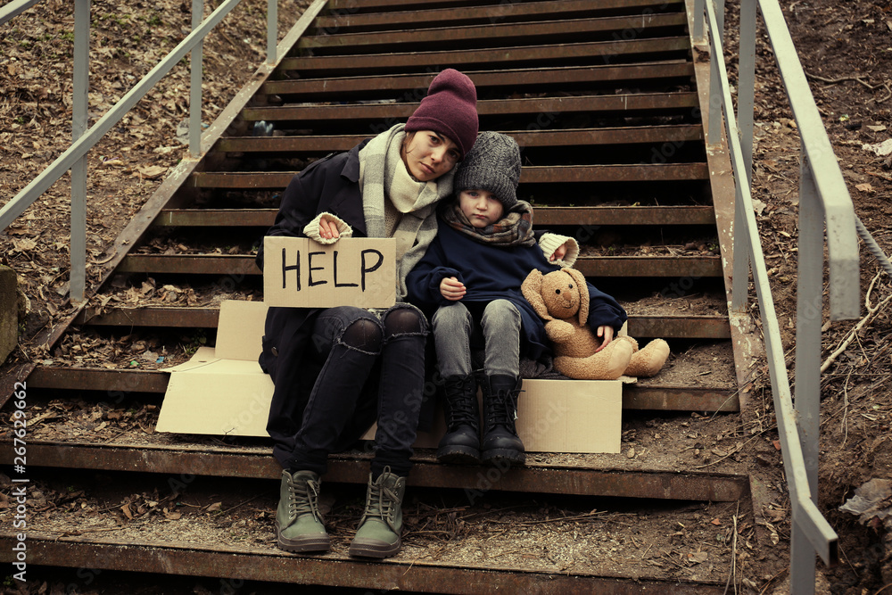 Poor mother and daughter with HELP sign sitting on stairs outdoors ...