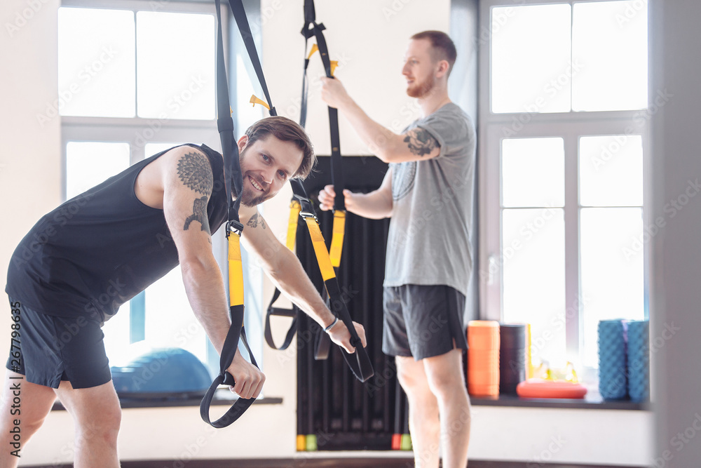 Positive young men athletes work out together in the gym using hanging ...