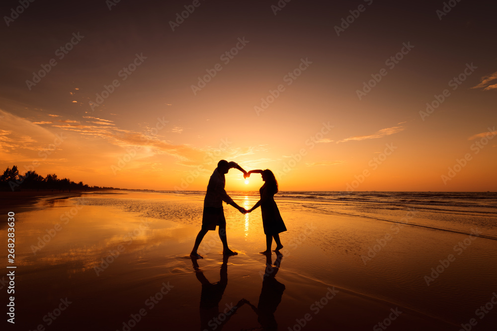 Silhouette of couple making heart shape with arms on beach at sunset ...