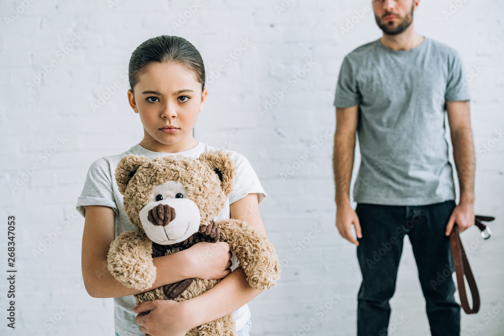 partial view of abusive father with belt and sad daughter holding teddy ...