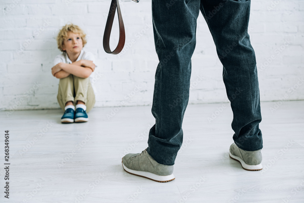 partial view of abusive father holding belt and son sitting on floor Photos | Adobe Stock