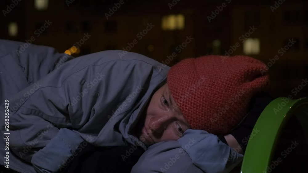sad homeless man cries when lying on a bench outdoors at night. Closeup ...