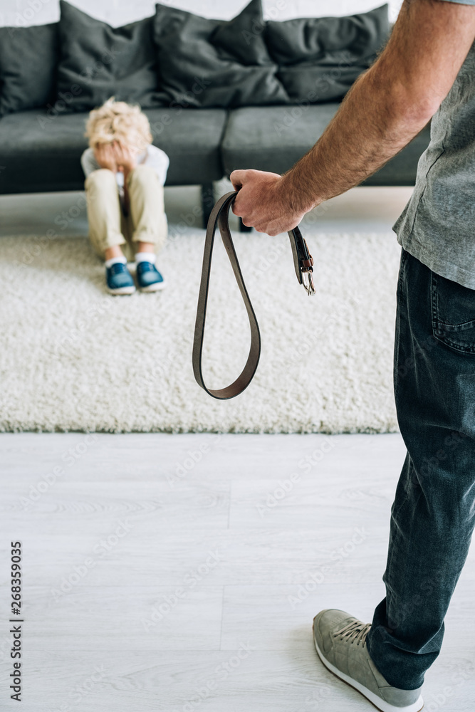 partial view of abusive father holding belt and sad son sitting on ...