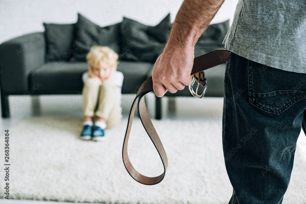 partial view of abusive father holding belt and sad son sitting on ...
