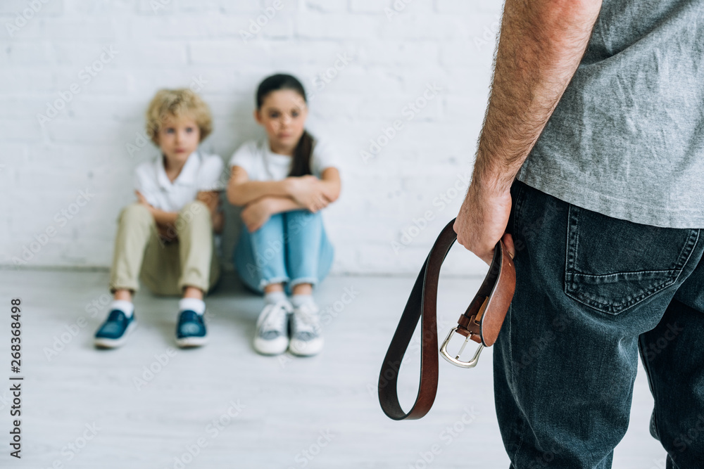 cropped view of abusive father holding belt and sad kids sitting on ...
