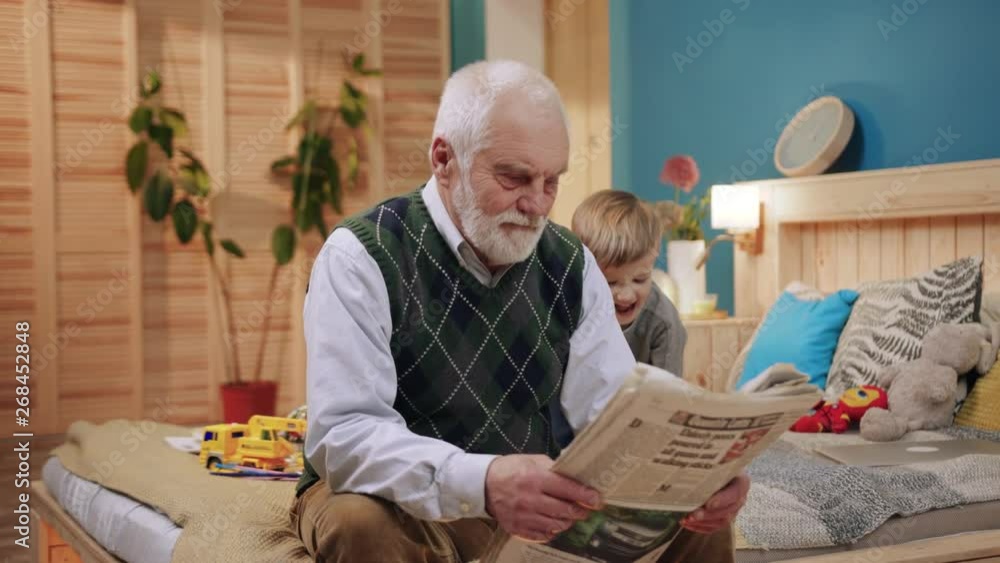 Old man in green vest sitting on bed with morning's newspaper in hands ...