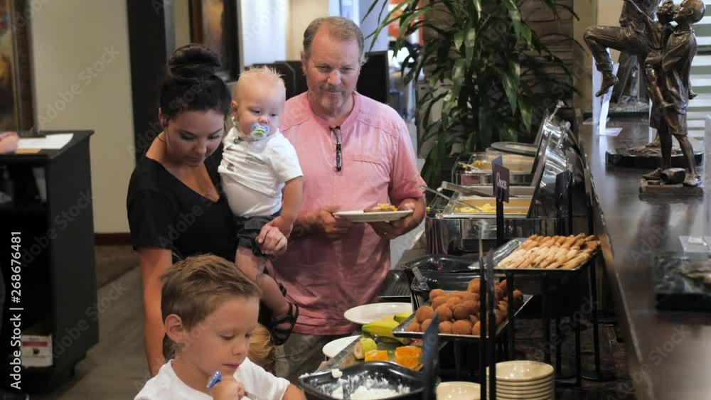 young mom dishes kids food from breakfast buffet line. A man waits in ...