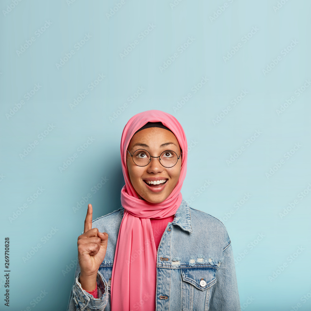 Vertical shot of happy dark skinned female Muslim woman poses over blue ...