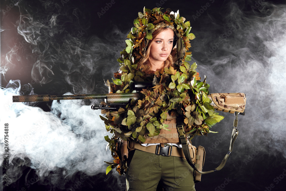 Camouflaged woman sniper in ghillie suit posing with rifle in foggy night. Special Weapons and ...
