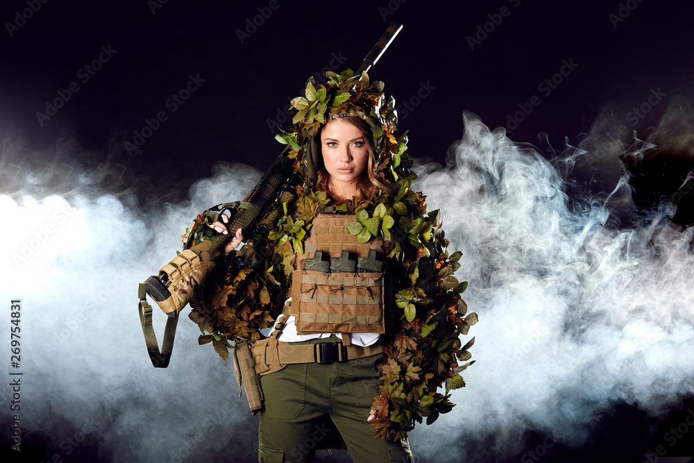 Camouflaged woman sniper in ghillie suit posing with rifle in foggy night. Special Weapons and ...
