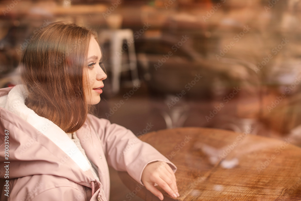 Beautiful young woman sitting at table in cafe, view from outdoors ...