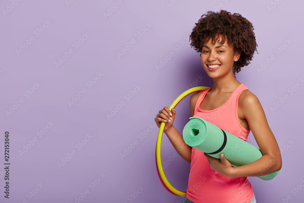 Delighted healthy woman rotates hoop, carries rolled karemat, has ...