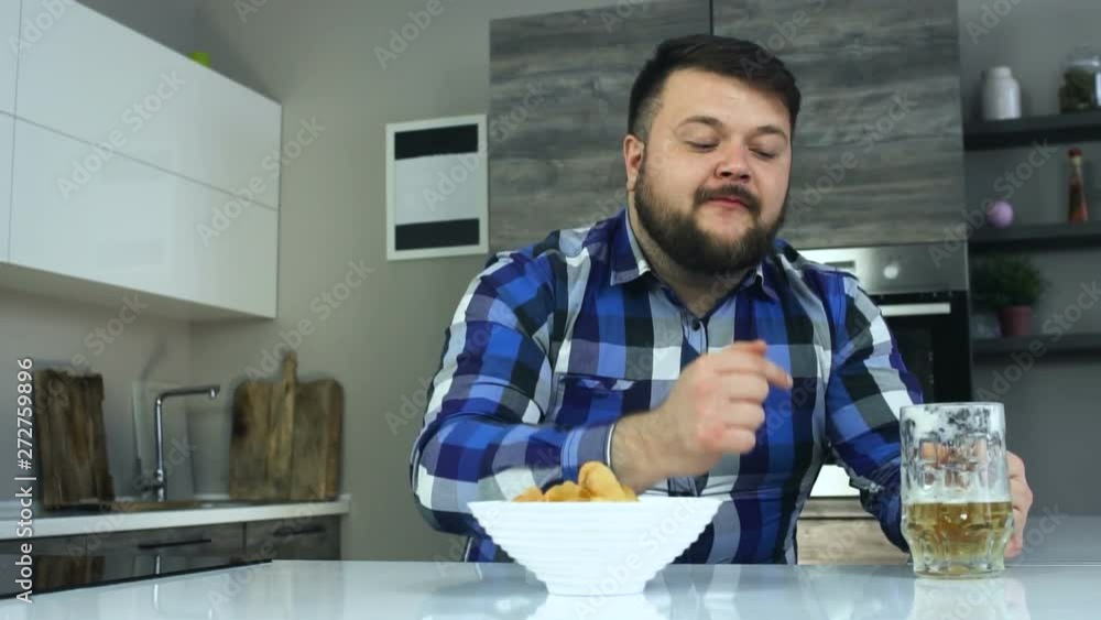 Fat man sits in kitchen at the table, eats crisps from bowl and drinks ...