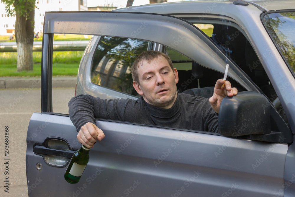 Drunk driving. Drunk driver hung on the door of his car with a bottle ...