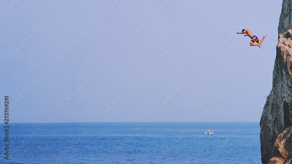 Young man jumping off cliff into blue water ocean at sunset. Active ...
