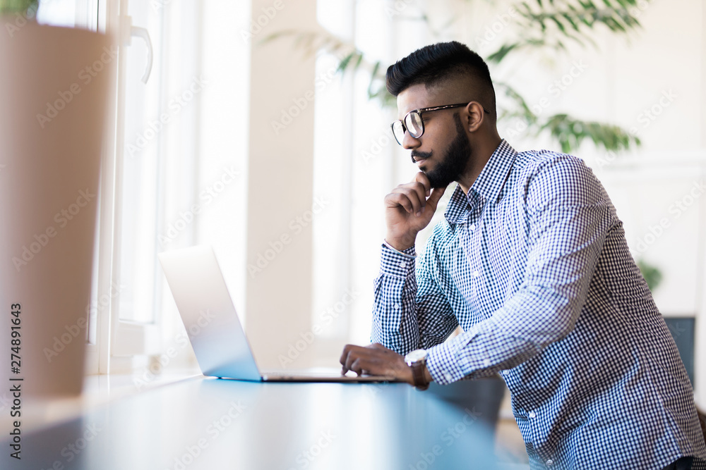 Indian young man with laptop standing in modern office background Stock ...